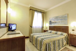 Quarto de hotel com cama, escrivaninha com computador, quadro na parede e janela com cortinas.