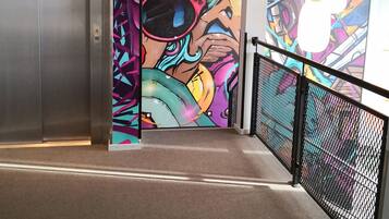 Um corredor interno com um mural colorido na parede, uma porta de elevador de metal e um guarda-corpo de metal preto.