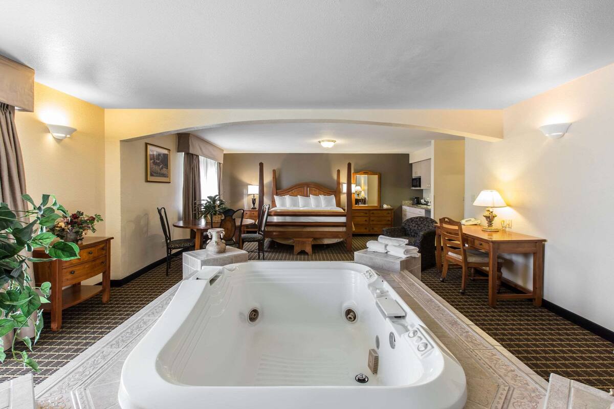 Quarto de hotel com uma banheira grande, uma cama com cabeceira de madeira, uma escrivaninha com luminária e uma cadeira.