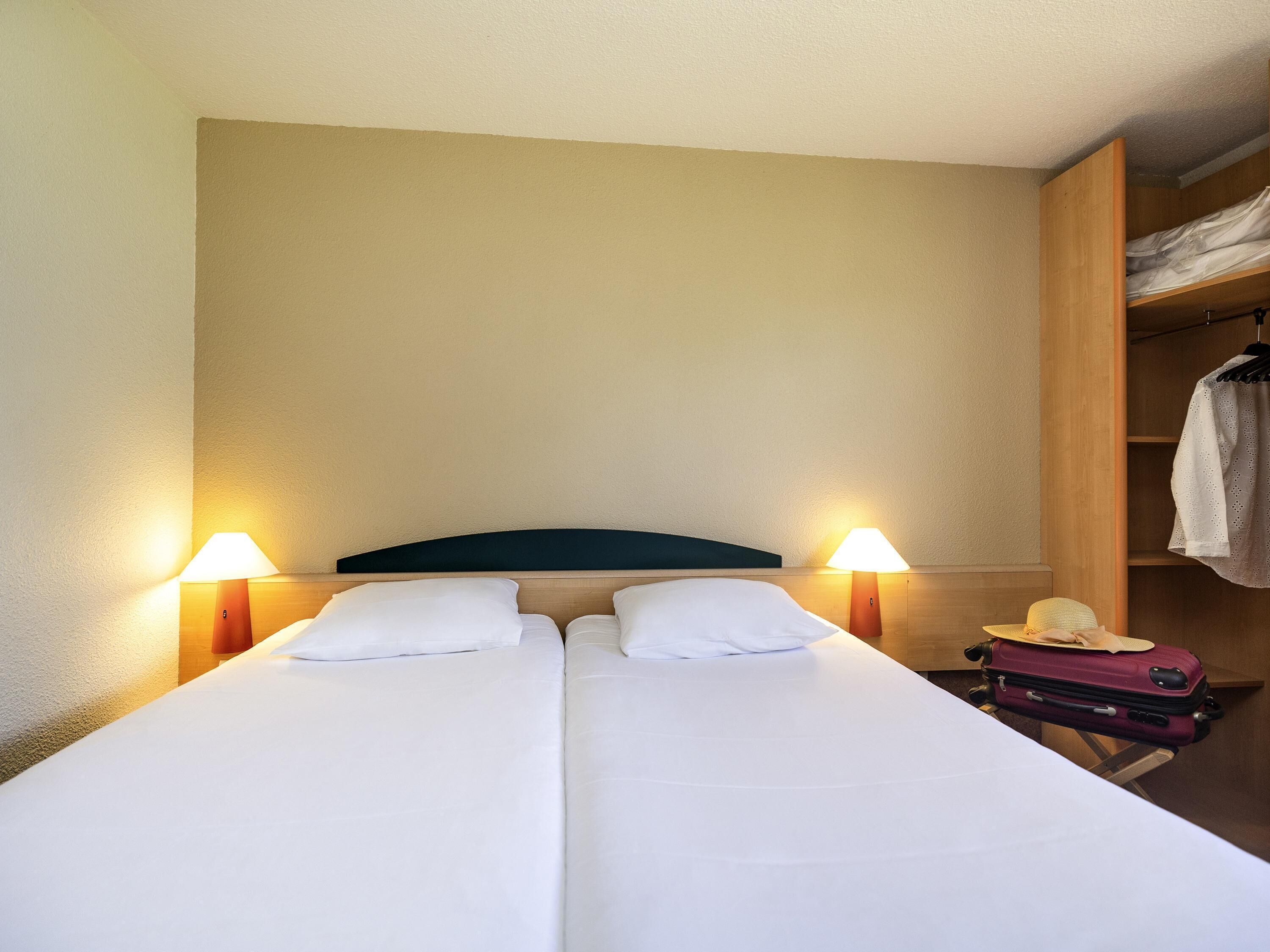 Uma cama de casal com duas luminárias de cabeceira, um painel de madeira e uma mala no chão.