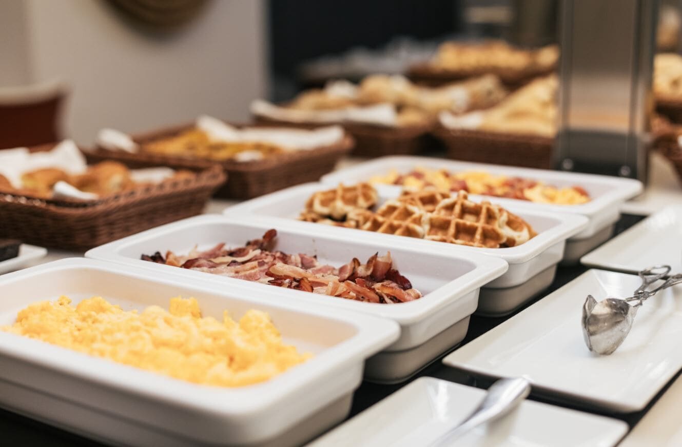 Buffet com diversas opções de comida, incluindo ovos mexidos, bacon, waffles e doces.