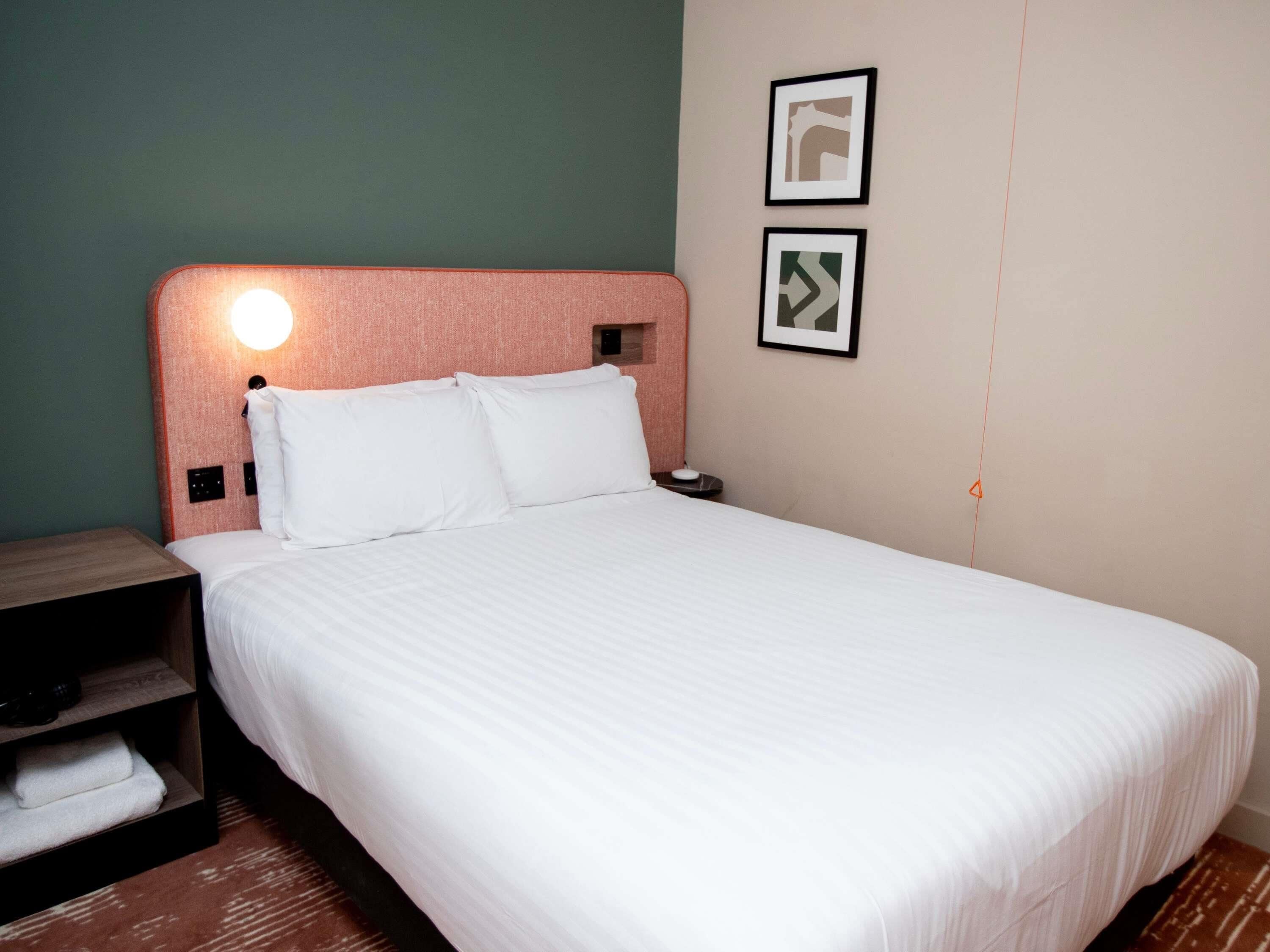 Uma cama bem arrumada com roupa de cama branca, uma mesa de cabeceira de madeira com uma luminária e duas fotos emolduradas na parede.