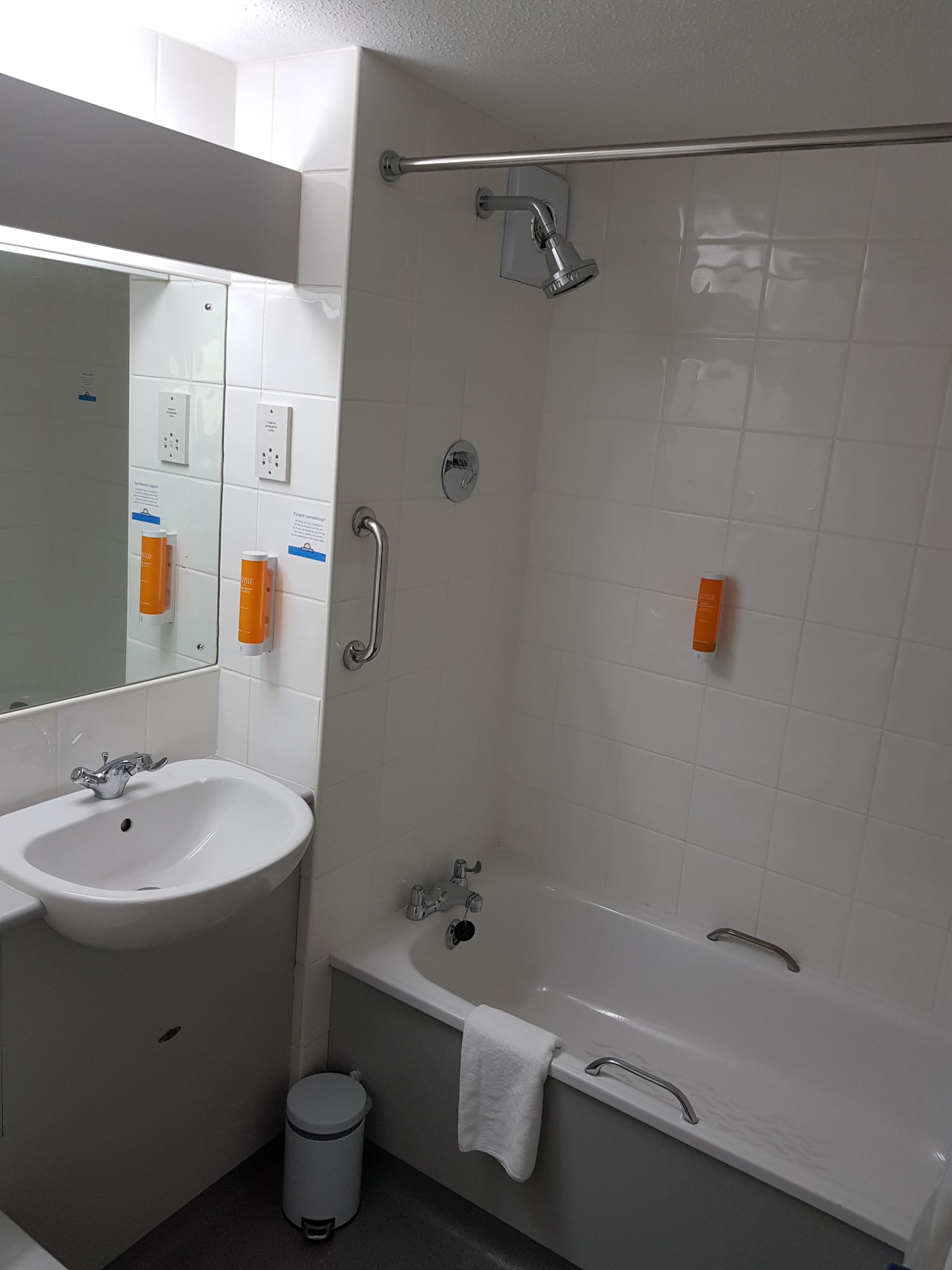 Casa de banho com lavatório branco, banheira, chuveiro com porta de vidro e barra de apoio.