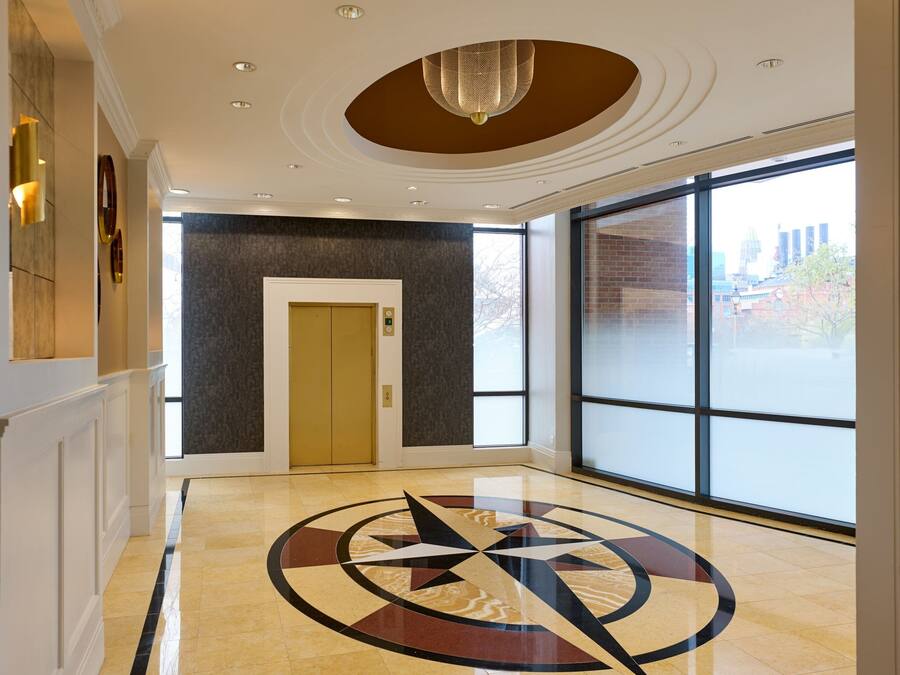 Um hall amplo com um grande design no chão, um elevador moderno e janelas grandes que oferecem vista para o exterior.