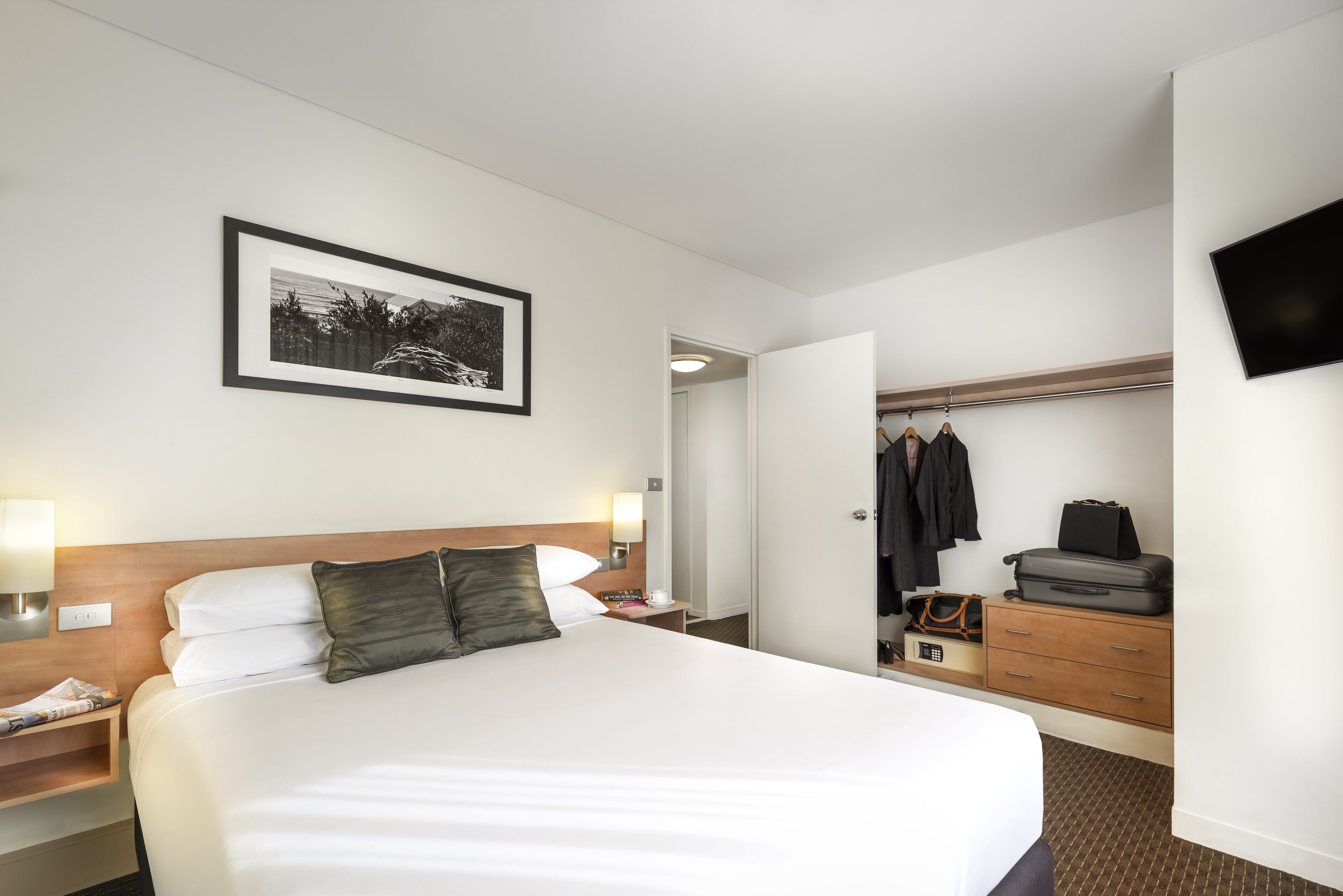 Quarto de hotel com uma cama grande, uma mesinha de cabeceira com abajur, um quadro na parede, um cabideiro com roupas e uma mala em um penteado.