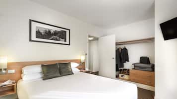 Quarto de hotel com uma cama grande, uma mesinha de cabeceira com abajur, um quadro na parede, um cabideiro com roupas e uma mala em um penteado.