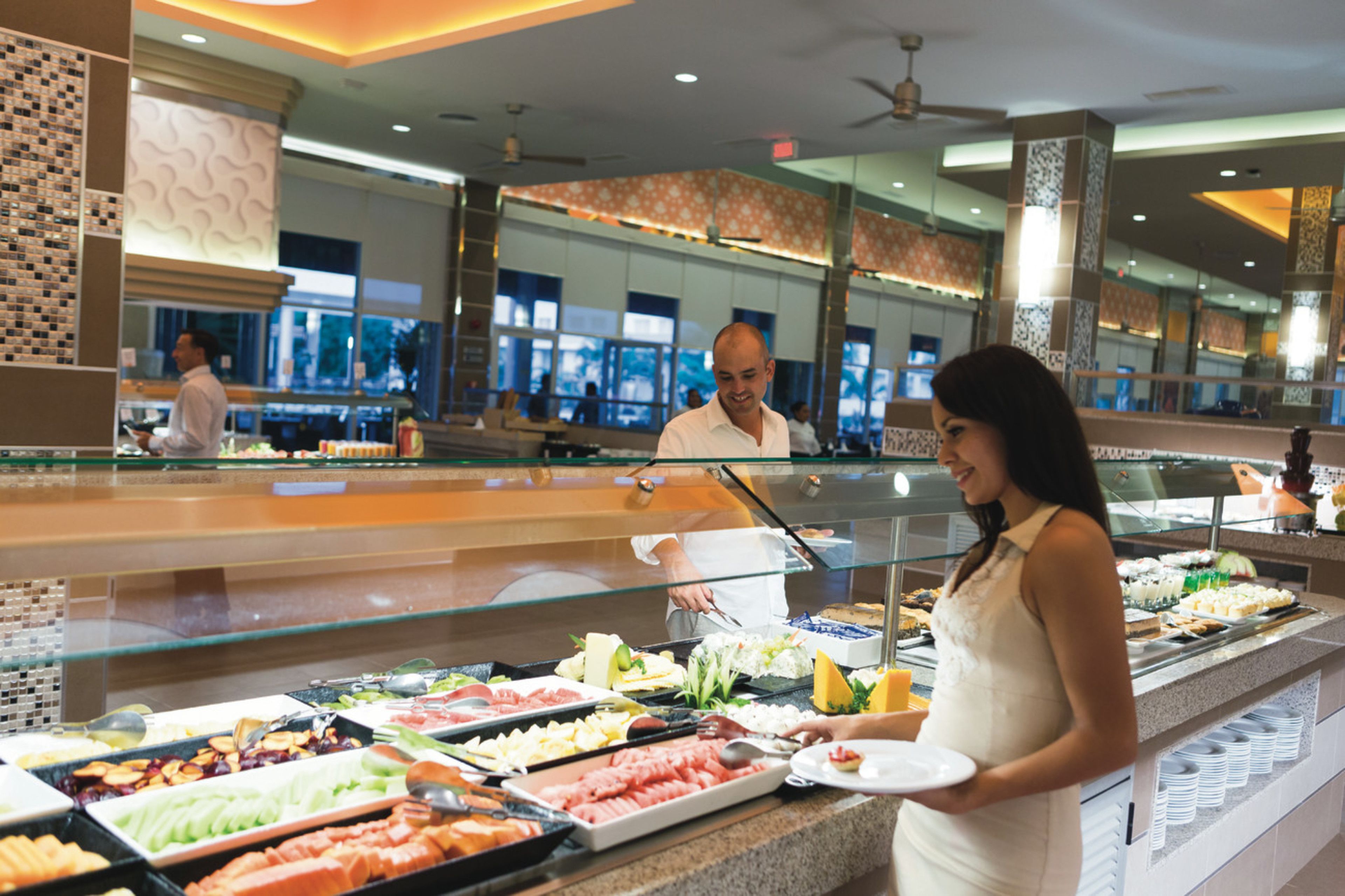 Espaço de buffet com diversas opções de comida, uma divisória de vidro e um funcionário servindo.