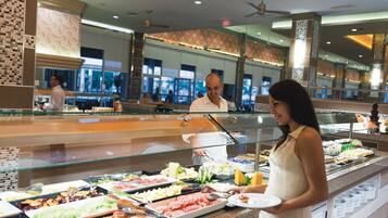 Espaço de buffet com diversas opções de comida, uma divisória de vidro e um funcionário servindo.