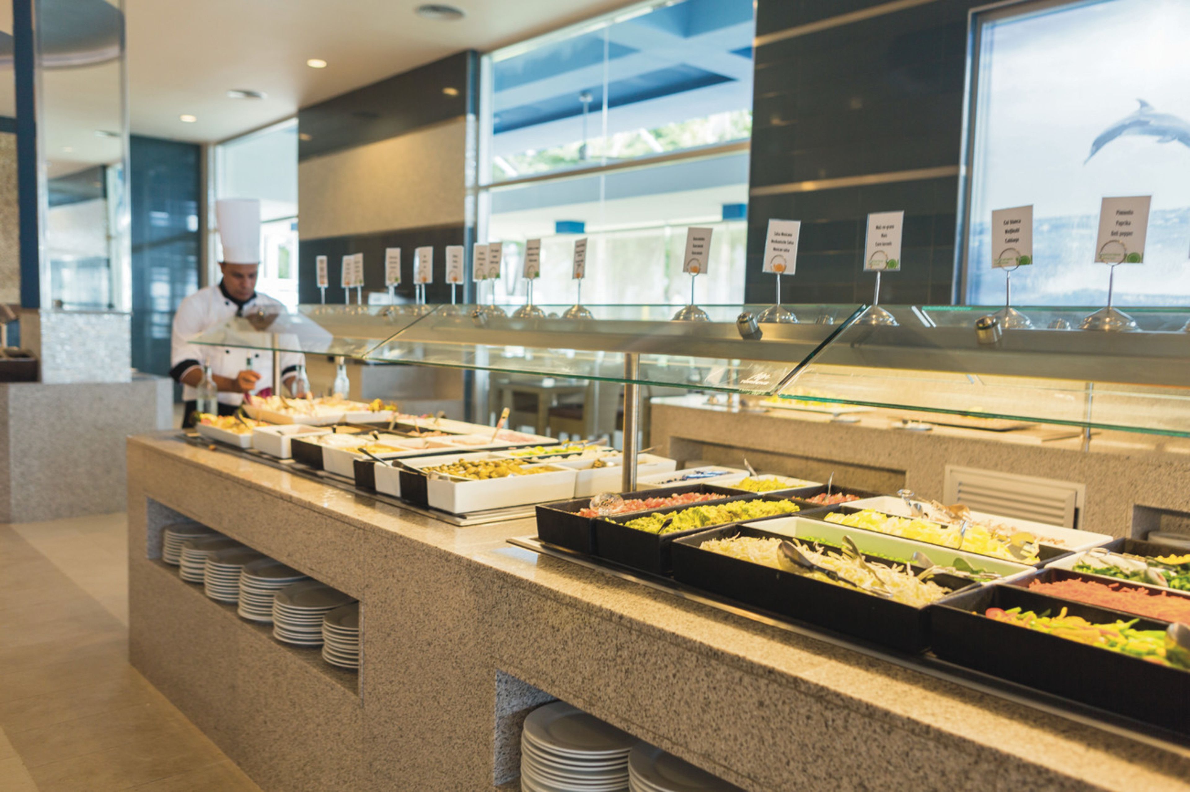 Buffet com diversas opções de comida, um chef uniformizado e vista para o mar através de amplas janelas.
