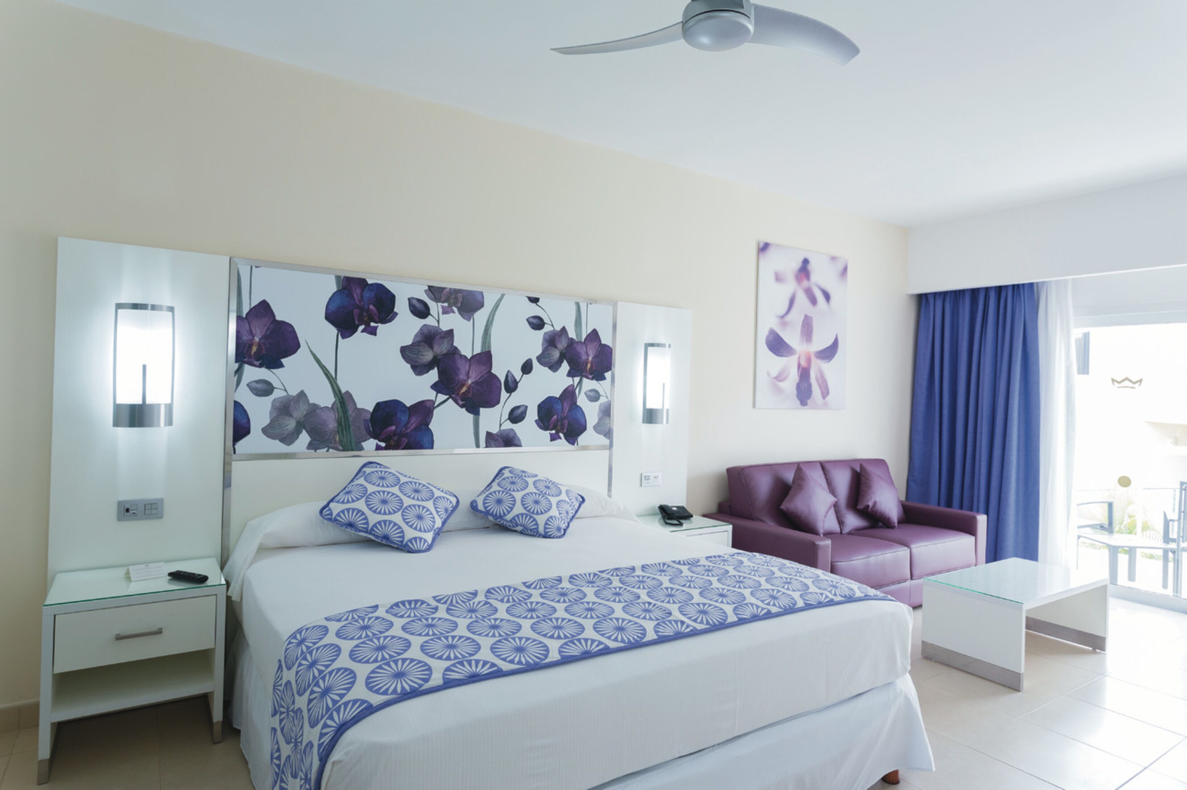 Quarto de hotel moderno com uma cama grande, um sofá roxo, uma mesa de centro de vidro e um ventilador de teto.