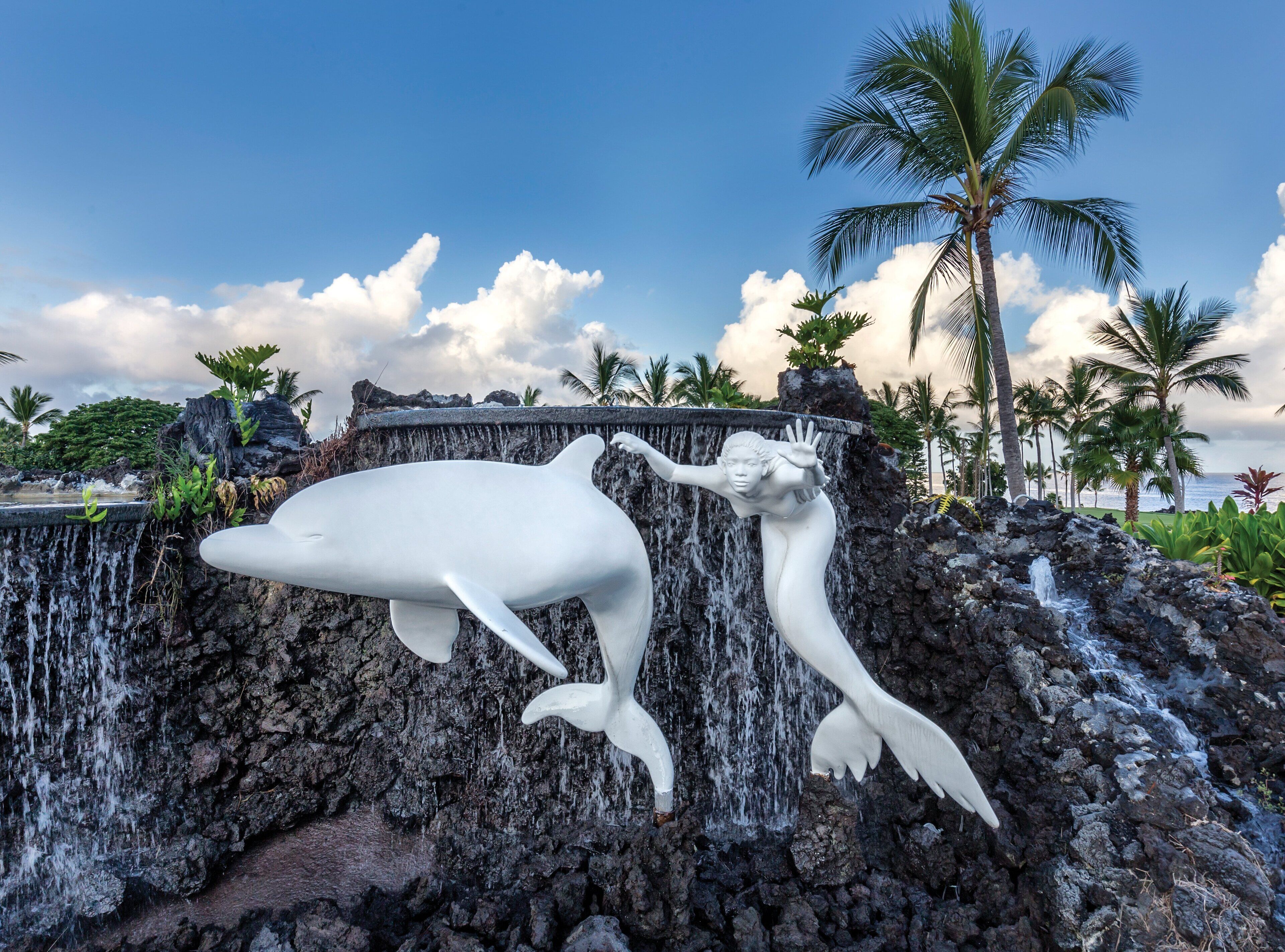 Escultura branca de um golfinho e uma sereia em um afloramento rochoso com uma cascata.