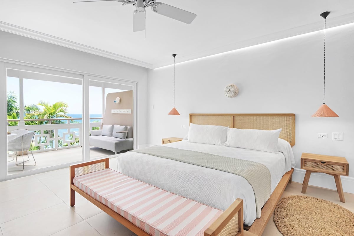 Quarto moderno com uma cama grande, um banco e vista para o oceano.