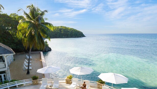 Uma área à beira da piscina com espreguiçadeiras e guarda-sóis, uma piscina e vista para o mar e uma ilha tropical.