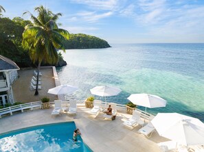 Uma área à beira da piscina com espreguiçadeiras e guarda-sóis, uma piscina e vista para o mar e uma ilha tropical.