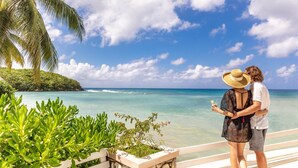 Um casal está em uma varanda com vista para uma praia tropical, com céu claro e ondas do oceano.