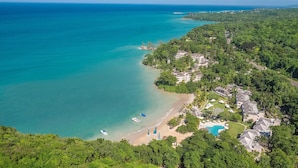 Vista aérea de um resort à beira-mar, com praia, piscina e vegetação exuberante.