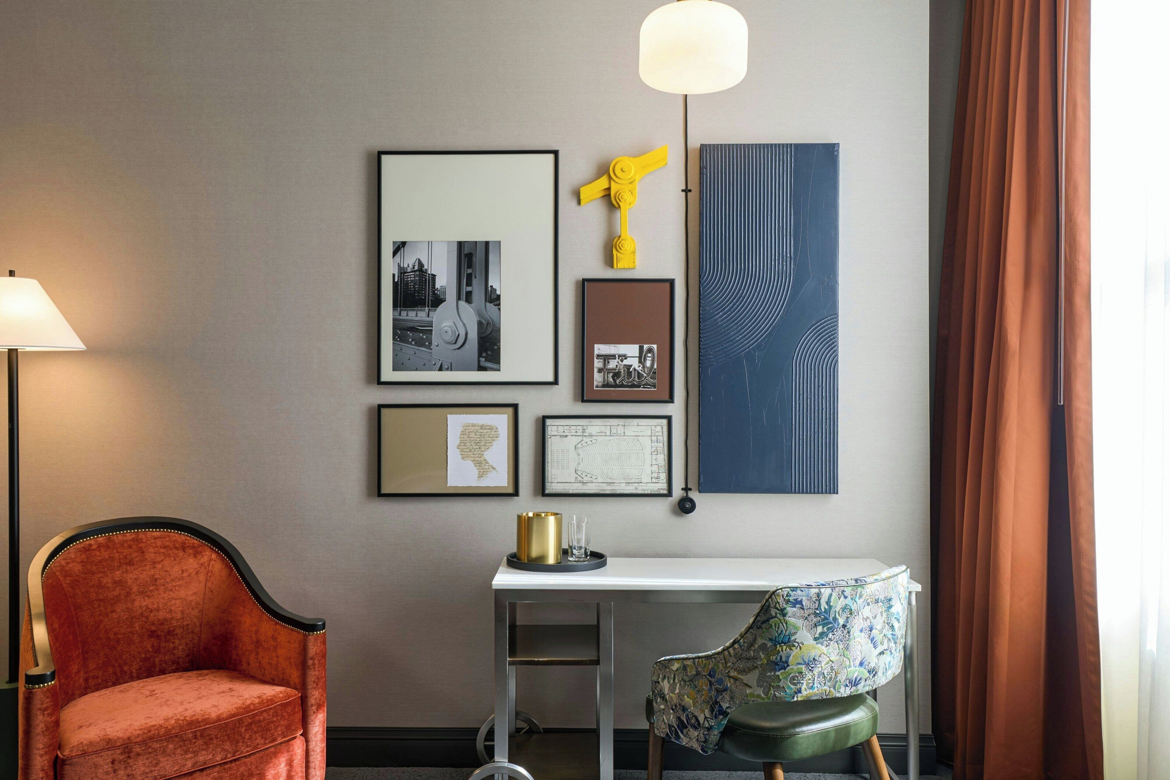 Quarto de hotel com uma poltrona de veludo vermelho, uma mesinha com abajur, e uma parede decorada com quadros e uma pintura abstrata azul.