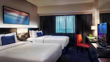 Quarto de hotel com duas camas, uma mesa de trabalho e uma televisão, além de uma janela grande com cortinas.