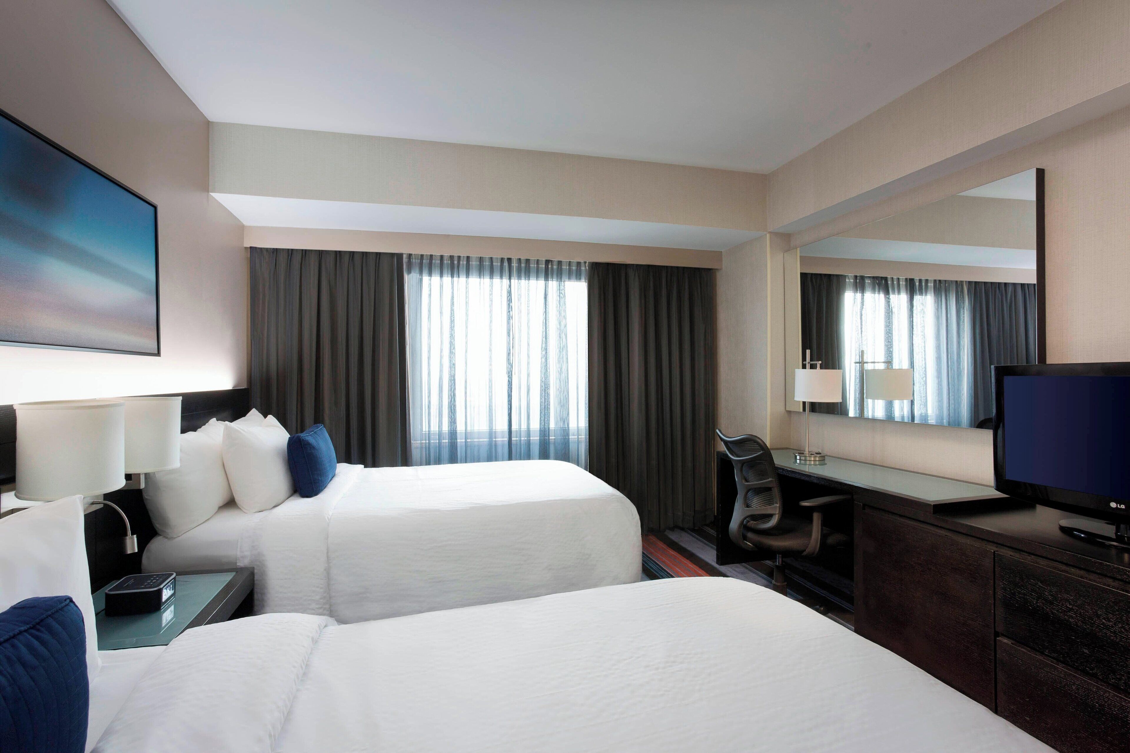 Quarto de hotel com duas camas, uma escrivaninha, uma televisão e um espelho grande.