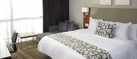 Quarto de hotel com cama grande, uma escrivaninha, uma cadeira, cortina escura e duas molduras na parede.