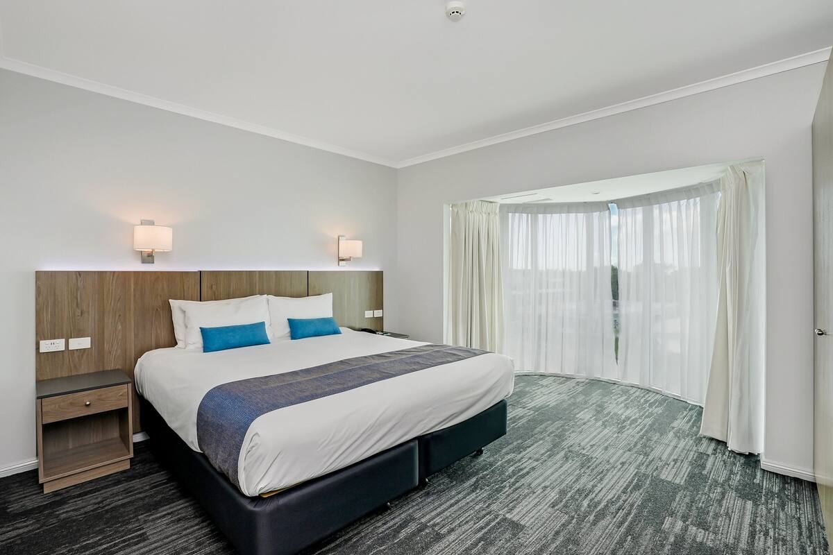 Quarto de hotel com uma cama grande, duas mesas de cabeceira, uma mesinha de apoio, uma janela com cortinas leves e vista para a cidade.
