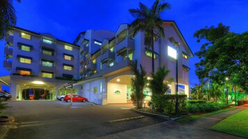 Um hotel de vários andares com design moderno, apresentando uma entrada central com passarela coberta e área de estacionamento com carros.