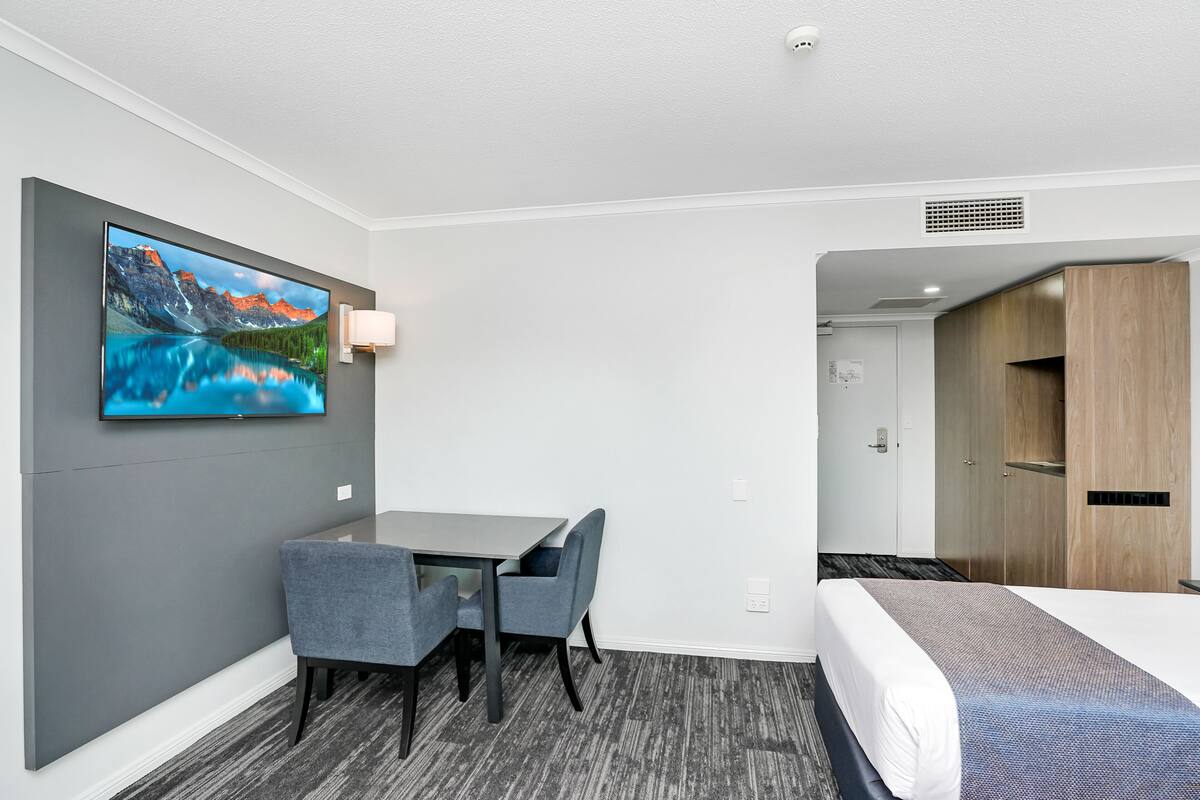 Quarto de hotel com uma cama, uma escrivaninha com duas cadeiras, uma televisão de tela plana na parede e um guarda-roupa de madeira.