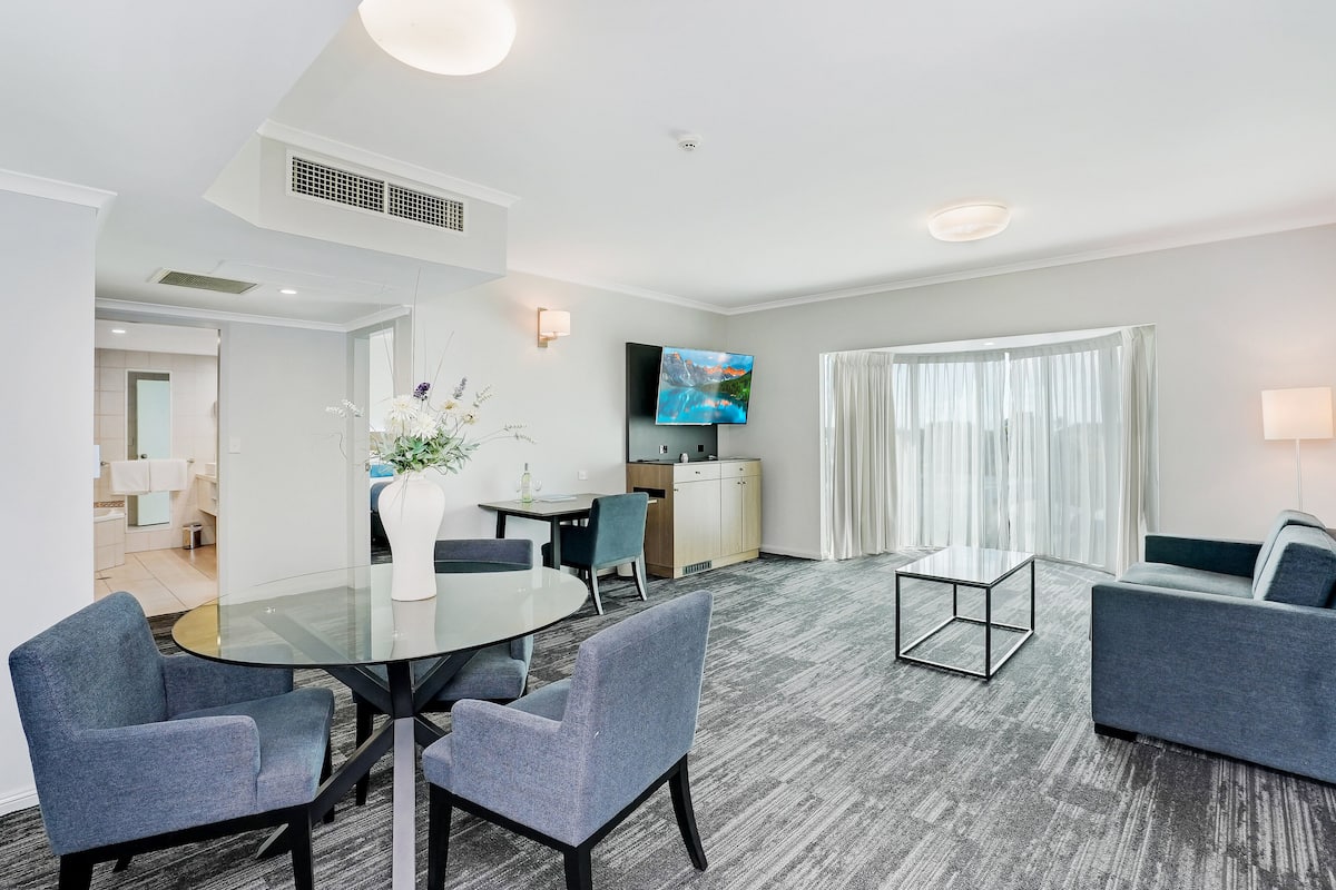 Quarto de hotel moderno com área de refeições, sofá, televisão e uma janela grande com cortinas.