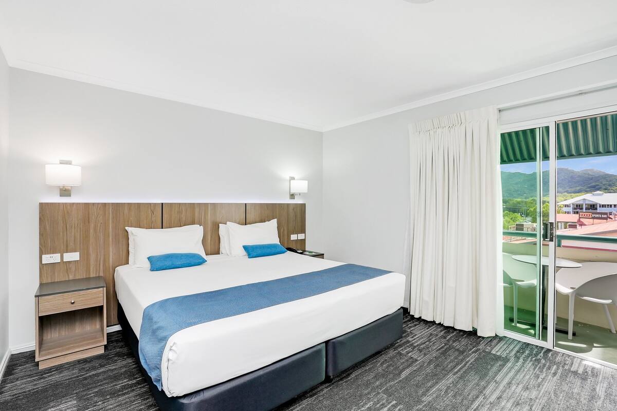 Quarto de hotel com uma cama grande, uma mesa de cabeceira, uma escrivaninha e vista para uma paisagem verde através de uma porta de correr de vidro.