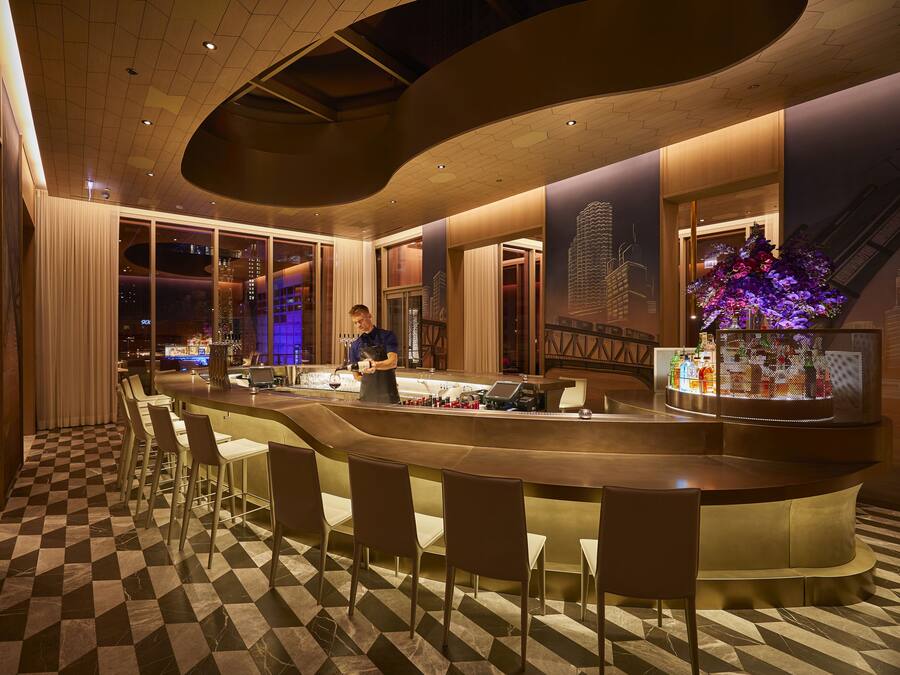 Uma área de bar moderna com chão quadriculado, banquetas e um barman.