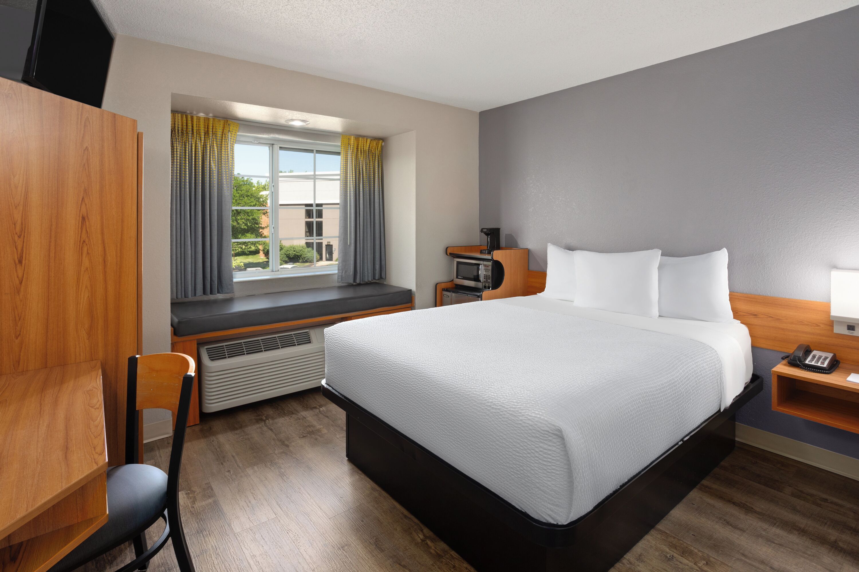 Quarto de hotel com cama, escrivaninha, cadeira, janela com cortinas, aquecedor, telefone e uma mesinha.