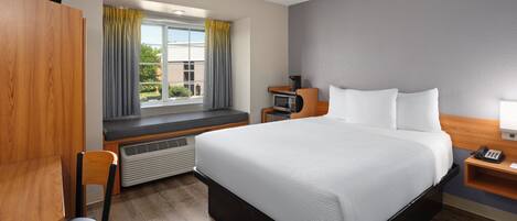 Quarto de hotel com cama, escrivaninha, cadeira, janela com cortinas, aquecedor, telefone e uma mesinha.