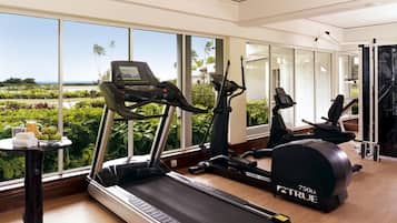 Uma academia com esteiras, elíptico e bicicleta ergométrica, com vista para uma paisagem tropical através de grandes janelas.