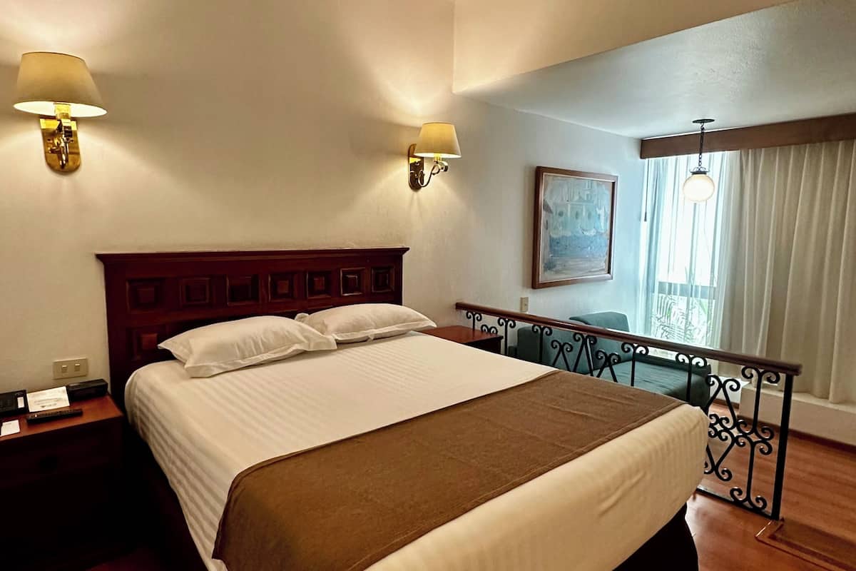 Quarto de hotel com uma cama grande, cabeceira de madeira, duas luminárias de cabeceira, mesa de cabeceira, janela com cortinas e um quadro na parede.