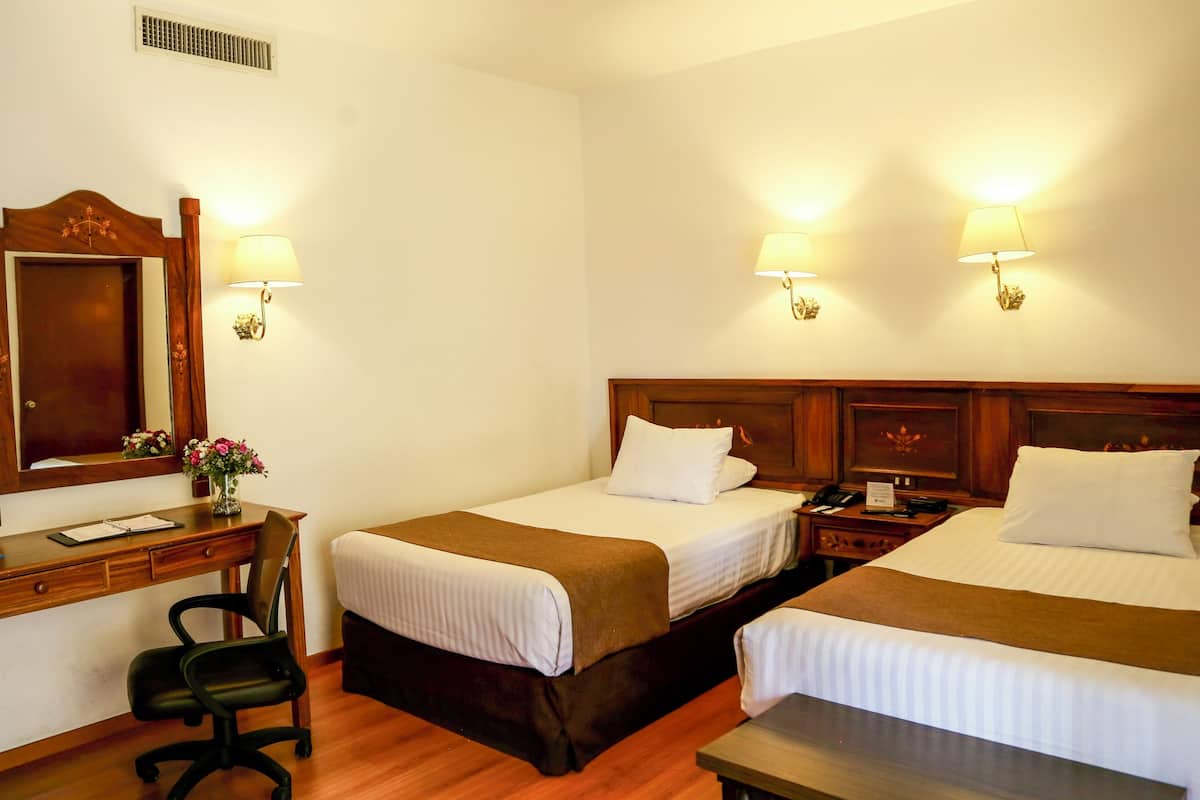 Quarto de hotel com duas camas de solteiro, uma escrivaninha com cadeira, um espelho e duas luminárias de cabeceira.