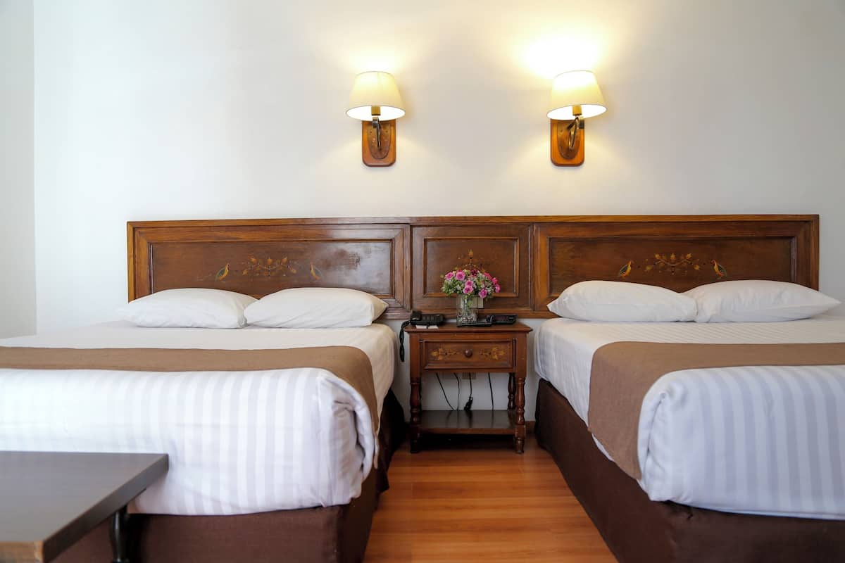 Quarto de hotel com duas camas, cabeceiras de madeira, mesas de cabeceira com luminárias e uma mesa de apoio com um vaso de flores.