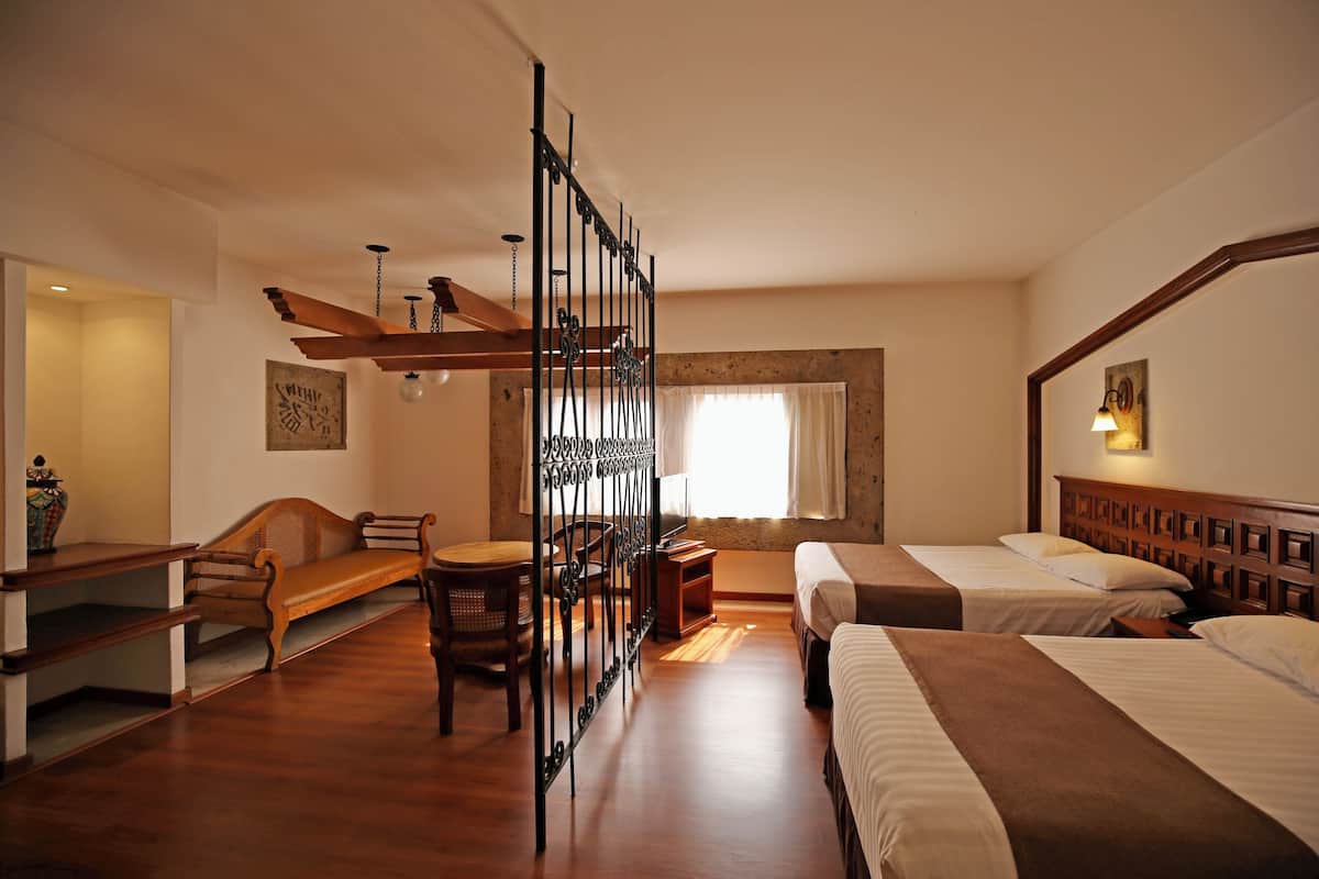 Quarto de hotel com um banco de madeira, duas camas de solteiro, uma escrivaninha com cadeira e um divisório metálico decorativo.