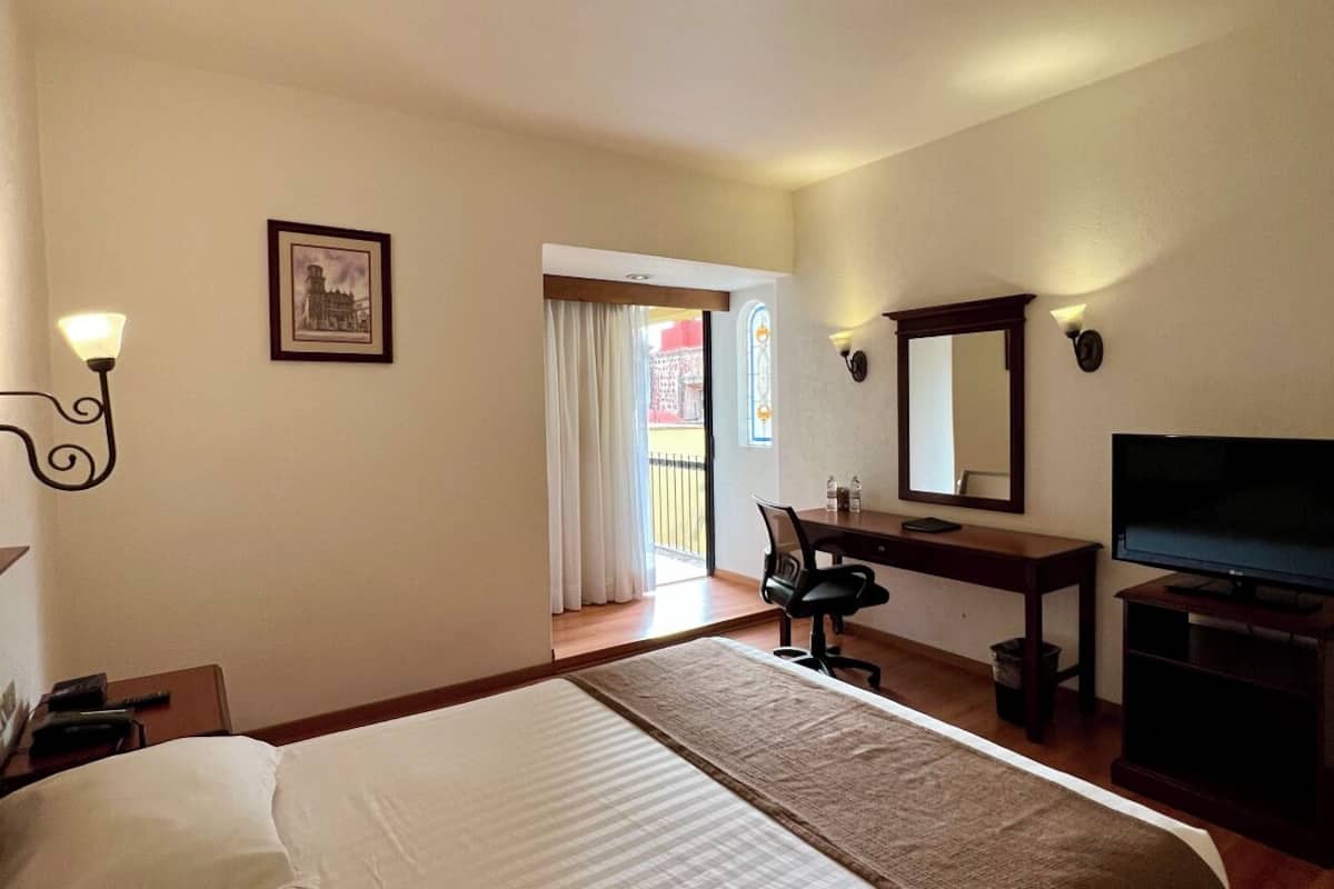 Quarto de hotel com uma cama grande, uma escrivaninha com cadeira, televisão e um quadro na parede.