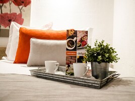Quarto de hotel bem arrumado com cama, almofadas, um suporte com xícaras e um vaso de planta.