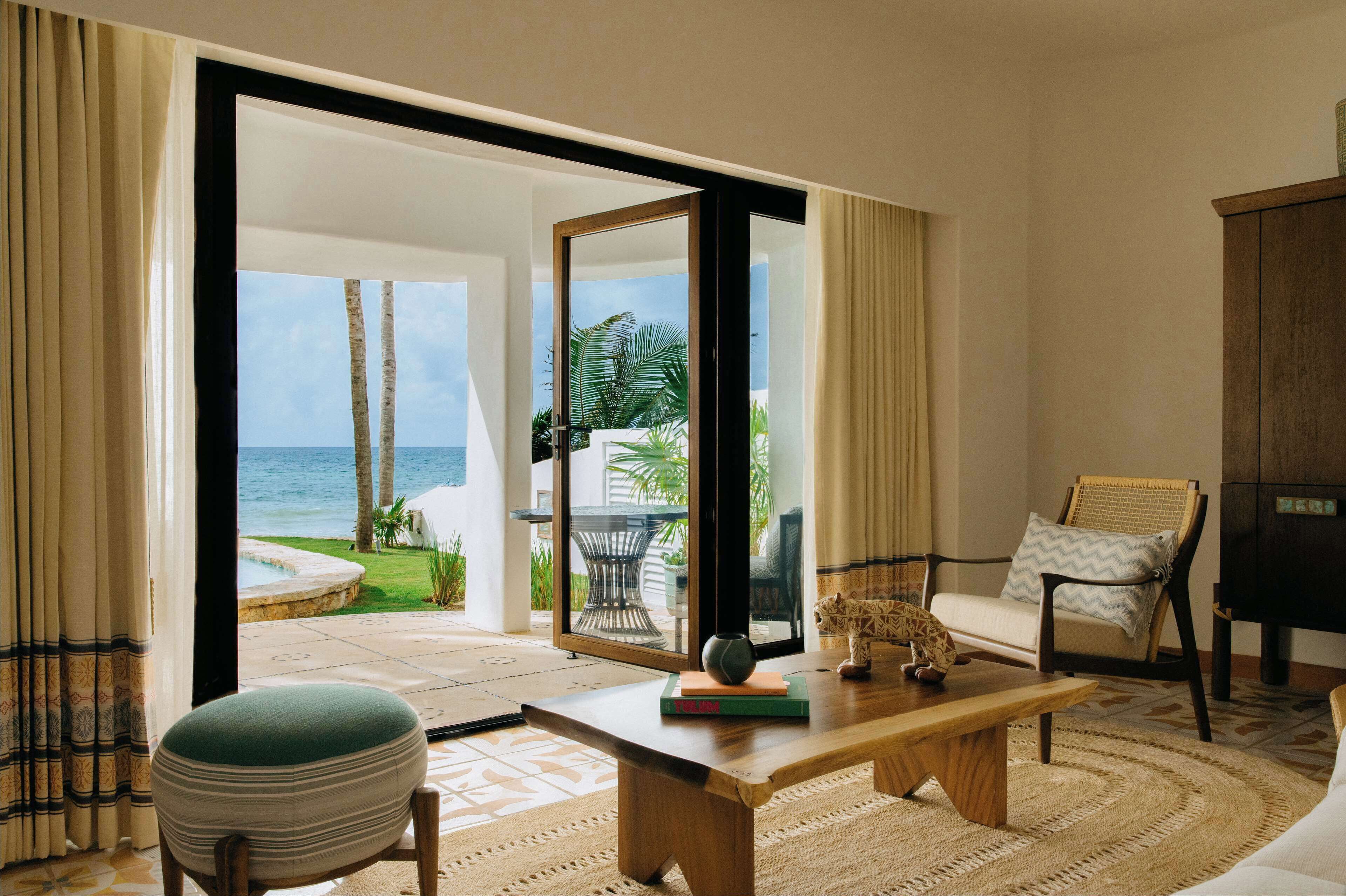 Quarto com uma grande porta de vidro que dá para uma varanda com vista para o oceano. Há uma mesa de madeira com livros e um objeto decorativo, uma cadeira de vime e um armário.