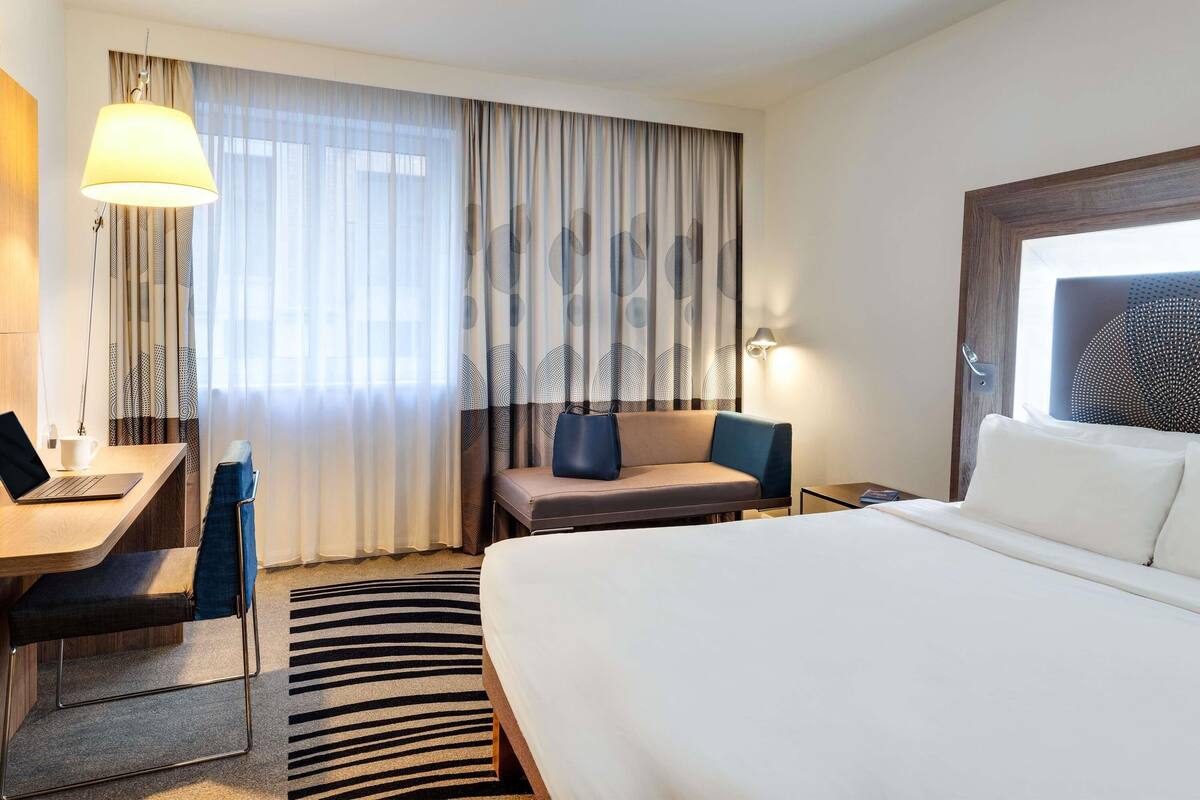 Quarto de hotel com cama, escrivaninha com laptop, cadeira, sofá e luminária.