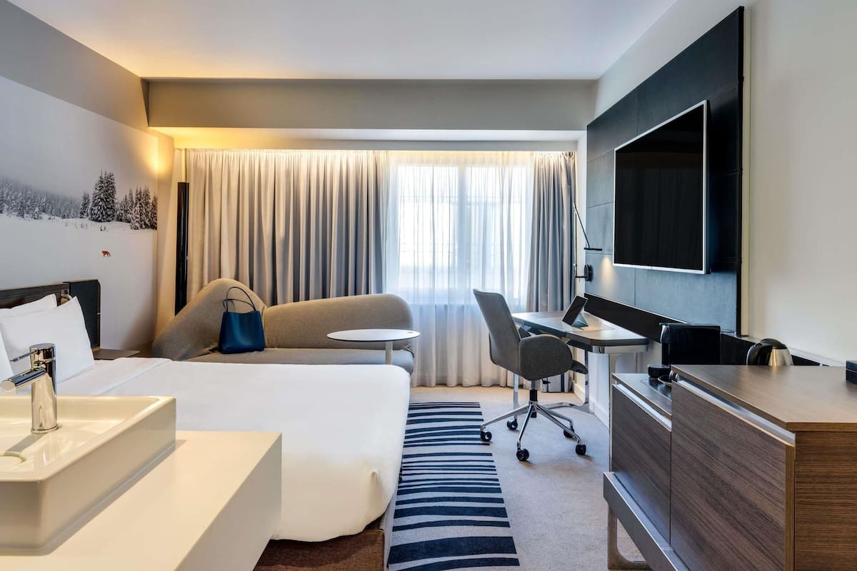 Quarto de hotel moderno com cama, escrivaninha, cadeira, televisão e uma grande janela com cortinas.