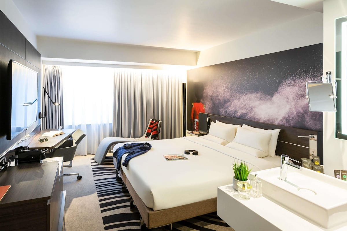 Quarto de hotel moderno com uma cama grande, uma escrivaninha, televisão, abajur, mesa de cabeceira, espelho e uma parede com um mural com tema de galáxia.