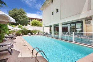 Piscina com espreguiçadeiras e guarda-sóis, cercada por um edifício com amplas janelas e céu claro.