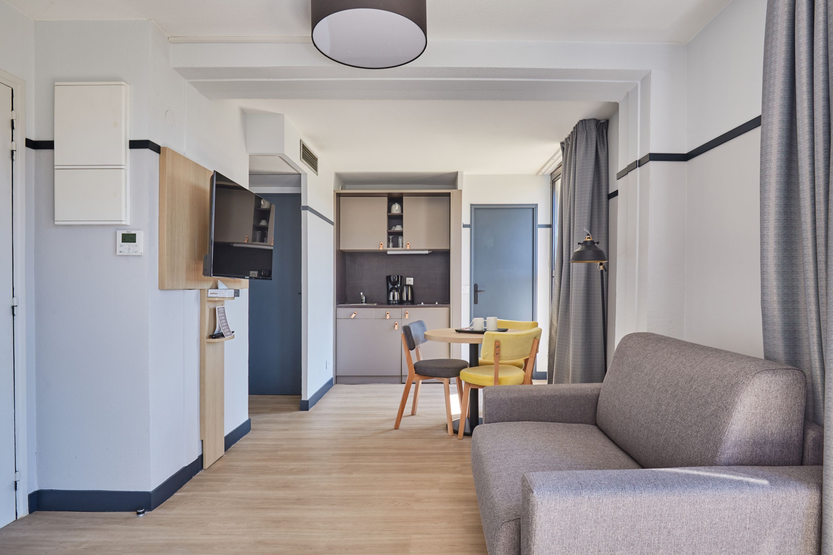 Quarto de hotel moderno com sofá, mesa de jantar com cadeiras, cozinha compacta e televisão na parede.