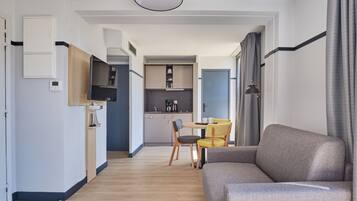 Quarto de hotel moderno com sofá, mesa de jantar com cadeiras, cozinha compacta e televisão na parede.