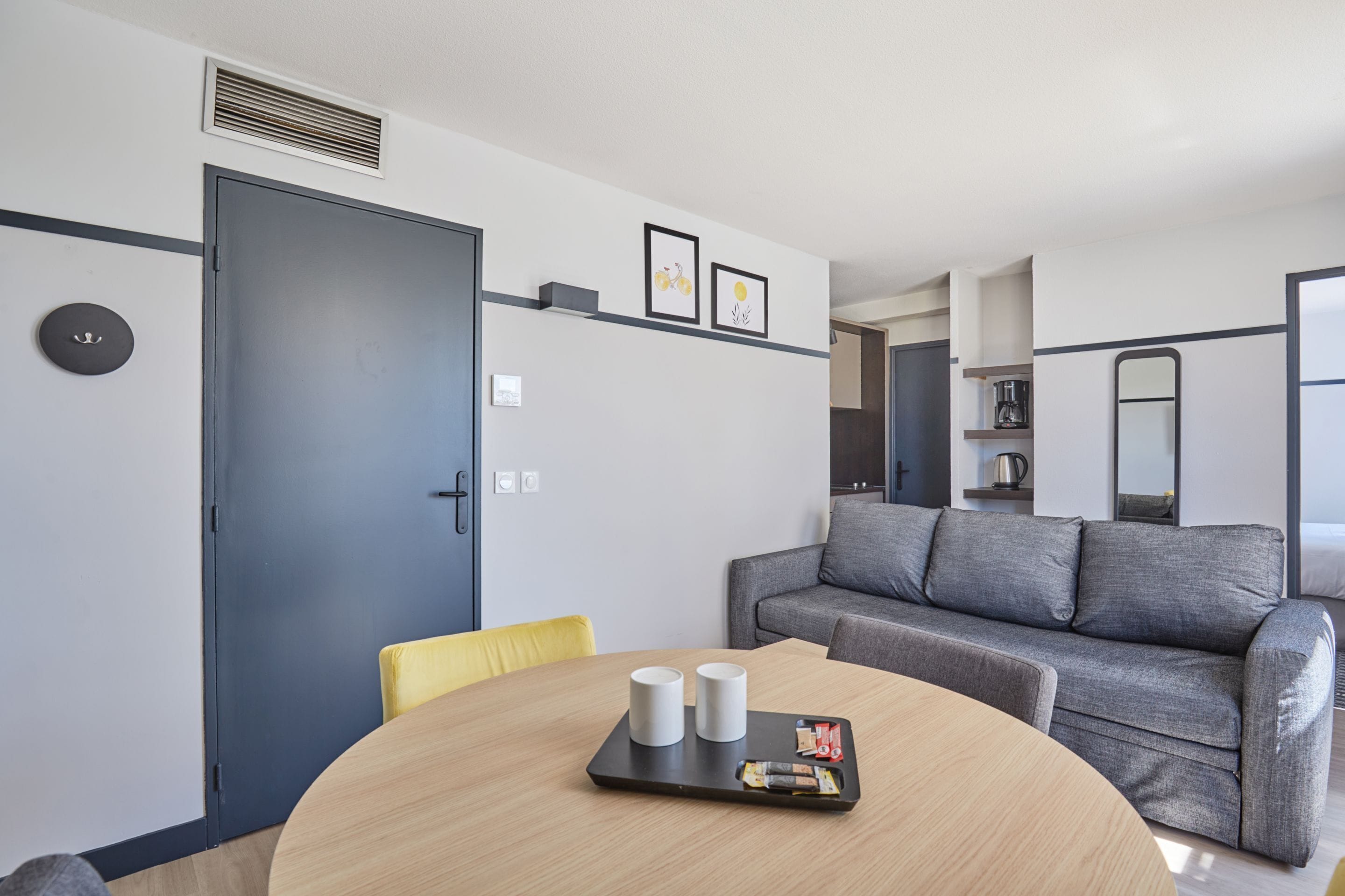 Quarto moderno com uma mesa redonda de madeira, duas chávenas brancas e um tabuleiro com um comboio de brincar. Um sofá cinzento e uma cadeira amarela estão visíveis.
