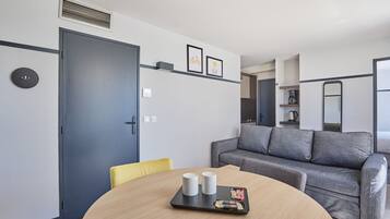 Quarto moderno com uma mesa redonda de madeira, duas chávenas brancas e um tabuleiro com um comboio de brincar. Um sofá cinzento e uma cadeira amarela estão visíveis.