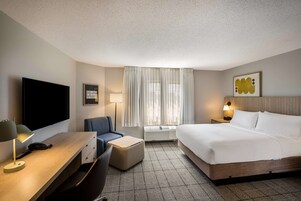 Quarto de hotel com cama, secretária com luminária, cadeira, televisão e janela com cortinas.