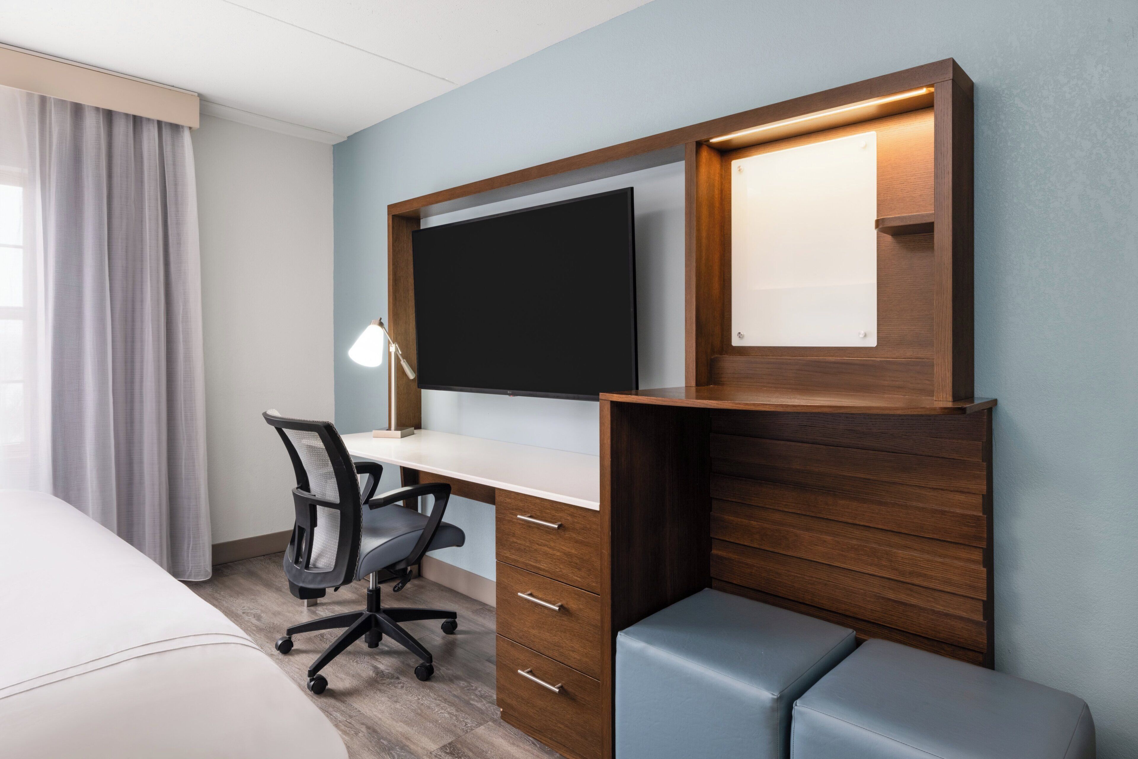 Quarto de hotel com uma cama, uma escrivaninha com cadeira, uma televisão e um pequeno banco.